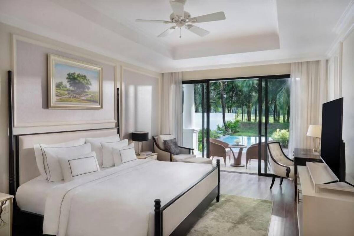 Quarto de hotel com uma cama grande, ventoinha no teto, televisão e varanda com vista para a piscina.