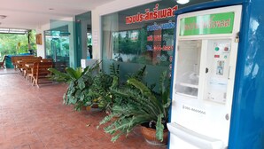Restaurante com mesas ao ar livre, máquina de vendas automática e placa com texto em tailandês.