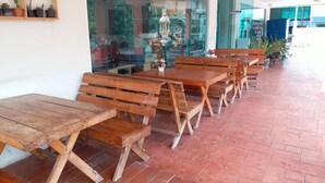 Uma área de pátio com mesas e bancos de madeira, uma porta de vidro que dá para o interior e plantas em vasos na parede.