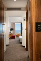 A hotel corridor with a wooden door labeled "303", a desk with a lamp, a red ottoman, and a view of the sea through a large window.