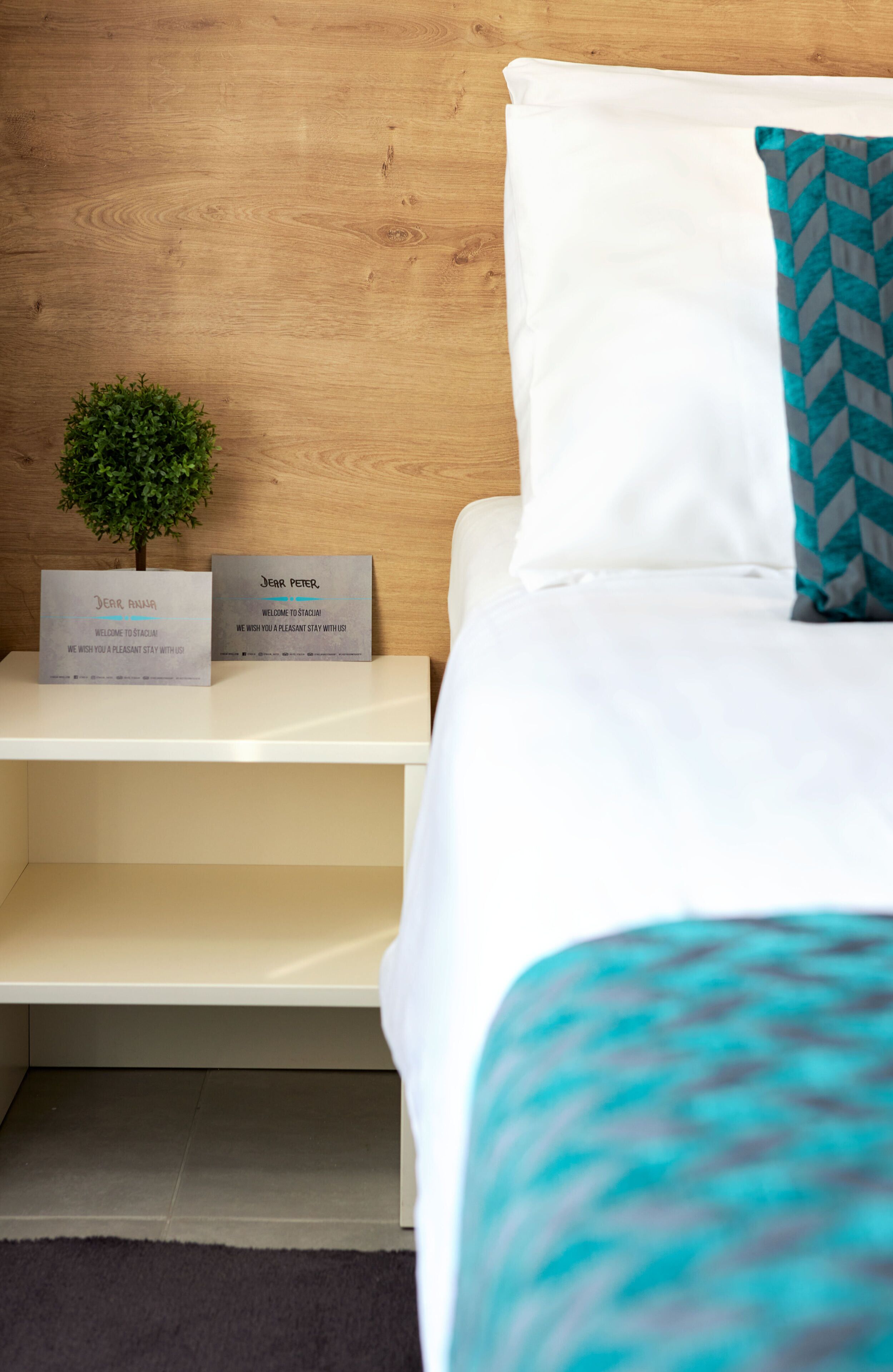 Cama bem arrumada com roupa de cama branca e verde-água, uma pequena planta em vaso e dois cartões de boas-vindas sobre uma mesa de cabeceira.