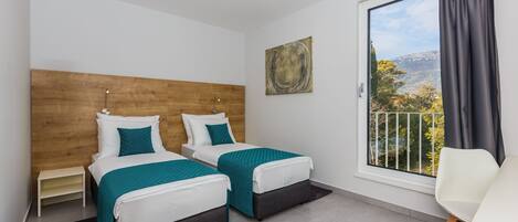 Quarto de hotel com duas camas, cabeceira de madeira, janela ampla com vista, uma cadeira branca e uma mesa pequena.