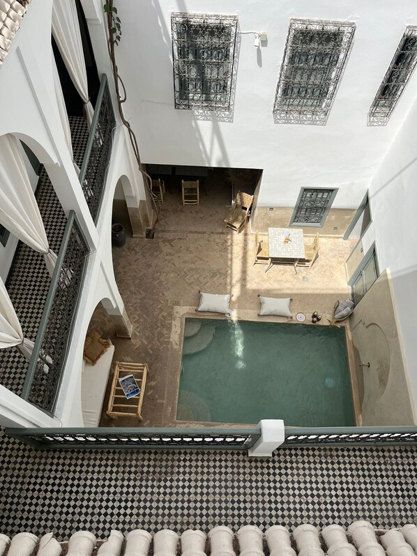 Uma área de piscina no terraço com piso xadrez, um espaço de estar pequeno com almofadas e vista para a arquitetura ao redor.
