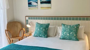Quarto de hotel com cama, duas almofadas, uma cadeira, mesa de cabeceira e duas obras de arte na parede.