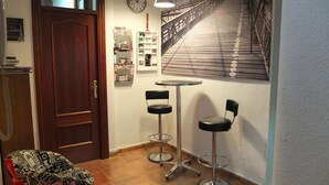 Um corredor com porta de madeira, uma cadeira estampada, dois bancos altos, uma mesa pequena e um relógio de parede.