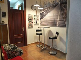 Um corredor com porta de madeira, uma cadeira estampada, dois bancos altos, uma mesa pequena e um relógio de parede.