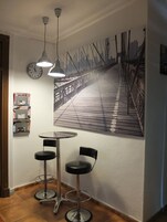 Uma área de refeições com uma mesa redonda e dois bancos altos, um relógio de parede e uma pintura com vista para a cidade.