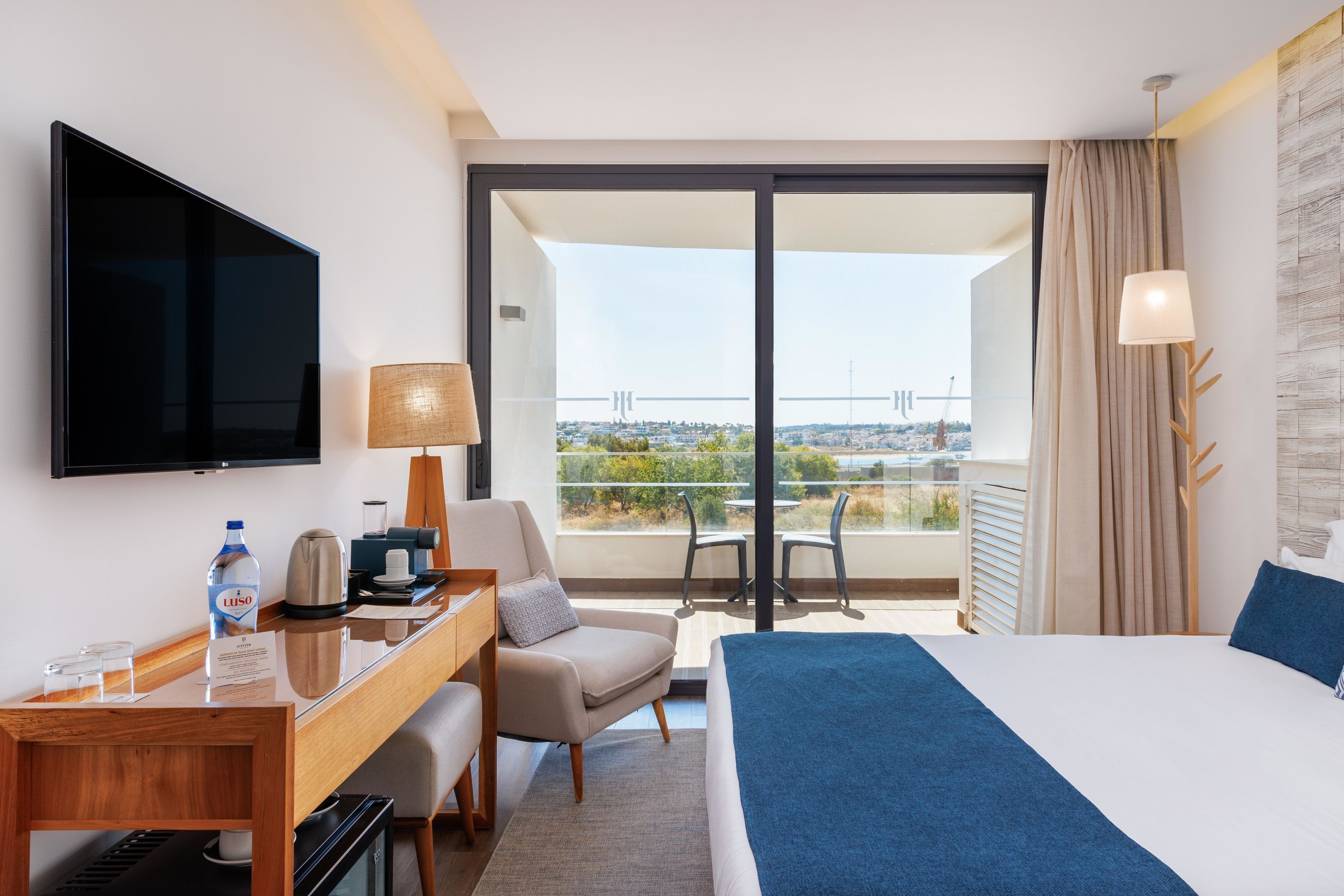 Quarto de hotel com uma cama, uma escrivaninha com abajur, uma televisão e vista da cidade através de amplas janelas.