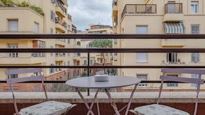 Varanda com mesa redonda e duas cadeiras, com vista para uma fileira de edifícios amarelos com varandas.