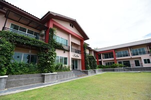 Um hotel de vários andares com fachada vermelha e bege, uma varanda no segundo andar e um gramado bem cuidado à frente.