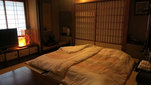 Um quarto tradicional japonês com piso de tatame, cama de madeira baixa com futon, televisão em suporte de madeira e um painel shoji atrás da cama.
