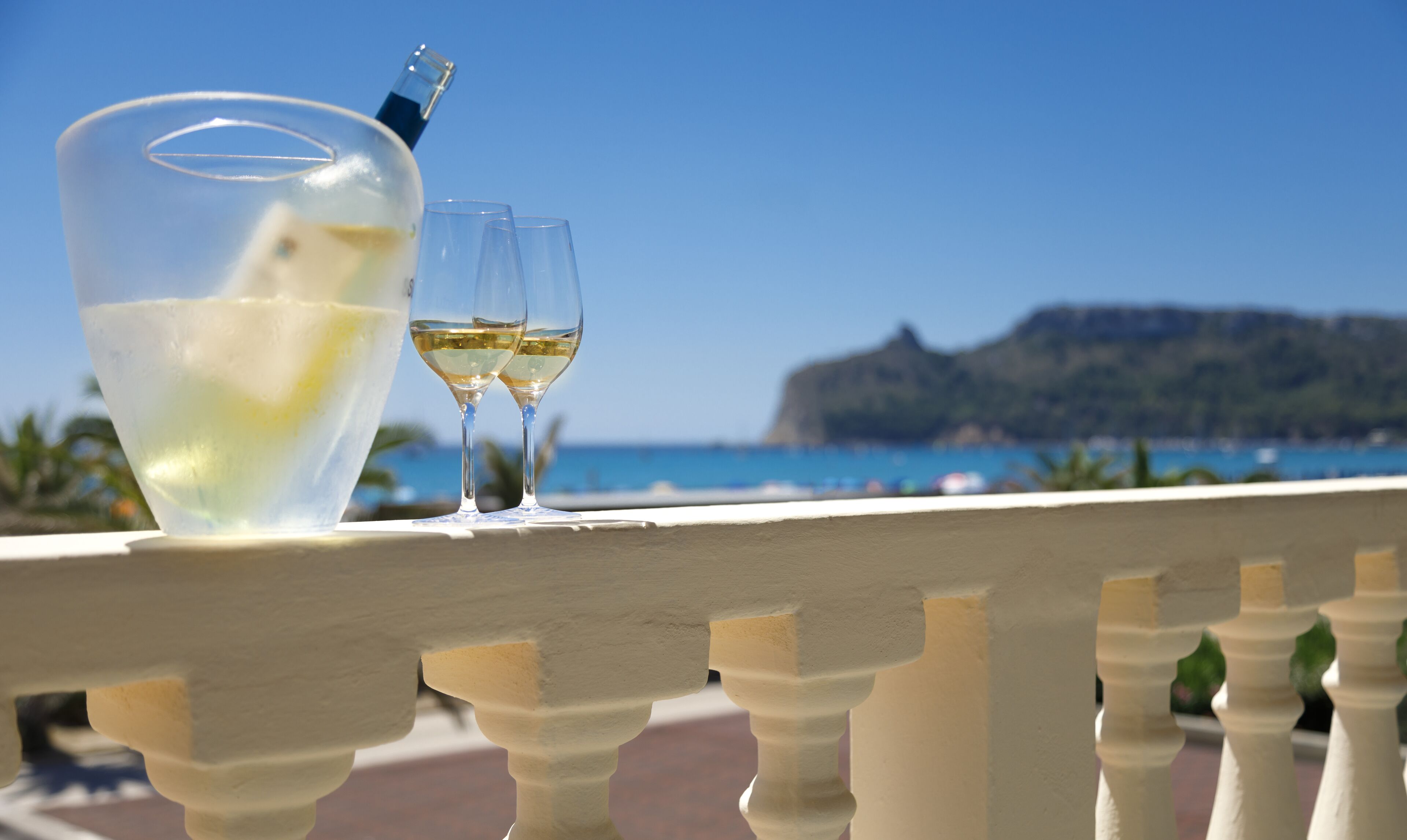 Um copo de vinho branco e dois copos de vinho numa varanda com vista para o mar e uma montanha ao fundo.