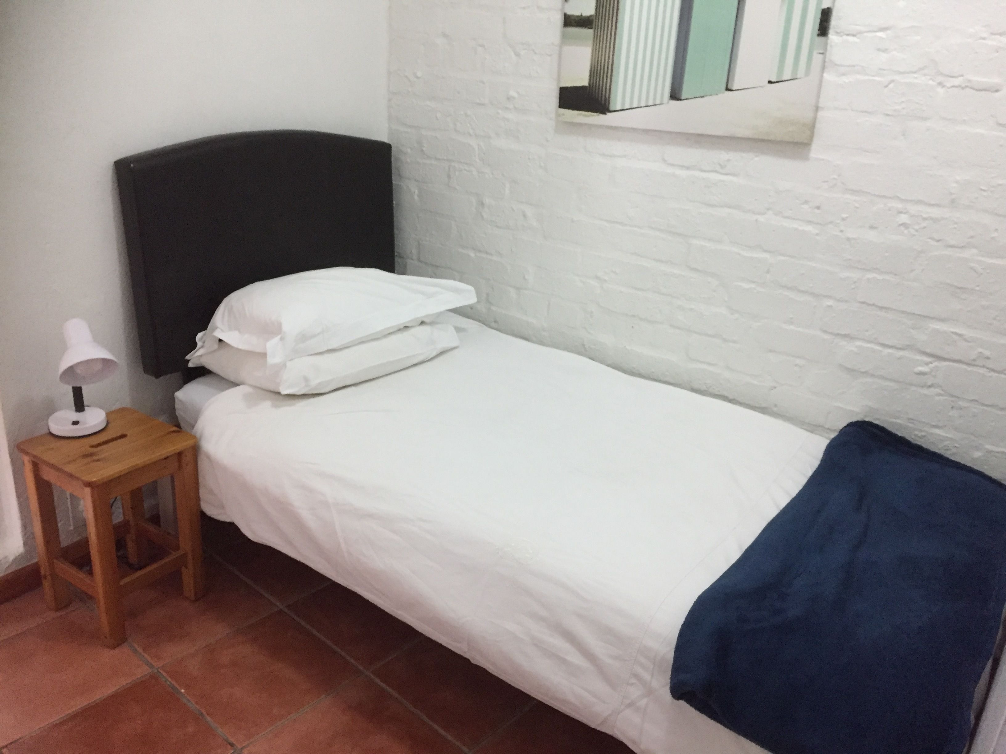 Uma cama pequena e bem arrumada com roupa de cama branca e um cobertor azul, uma mesinha de cabeceira de madeira com uma luminária branca, e uma parede de tijolos brancos com uma janela acima da cama.