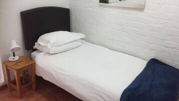 Uma cama pequena e bem arrumada com roupa de cama branca e um cobertor azul, uma mesinha de cabeceira de madeira com uma luminária branca, e uma parede de tijolos brancos com uma janela acima da cama.