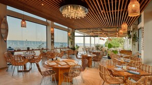 Um restaurante espaçoso com mobiliário de vime, amplas janelas com vista para o mar e teto de madeira com luminárias pendentes.