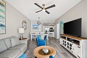 Uma sala de estar com um sofá cinzento, uma poltrona azul, um espaço de jantar com cadeiras brancas, uma cozinha ao fundo e uma TV sobre um móvel de madeira.