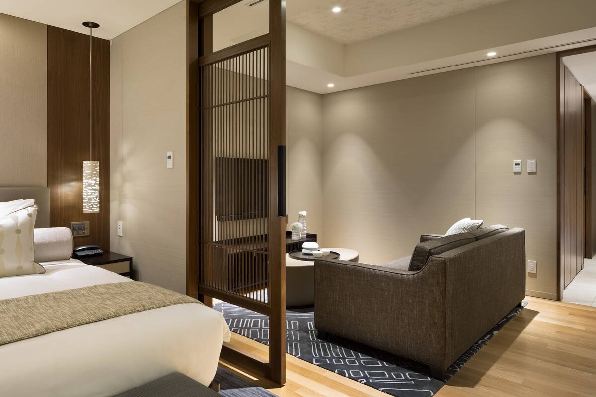Quarto de hotel moderno com uma cama, um sofá, um painel de madeira e uma pequena mesa com abajur.