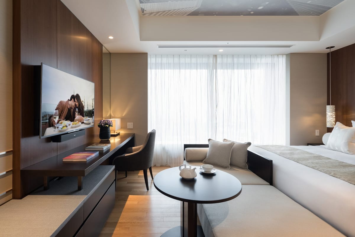 Quarto de hotel moderno com uma cama grande, um sofá, uma pequena mesa redonda com um bule e uma televisão de tela plana montada na parede, além de uma janela grande com cortinas leves.