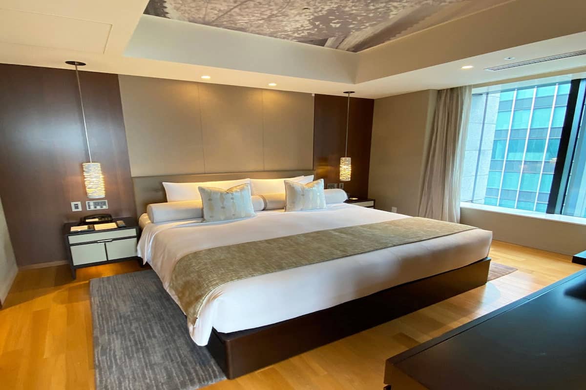 Quarto de hotel moderno com uma cama grande, mesas de cabeceira, uma escrivaninha e vista para a cidade pela janela.