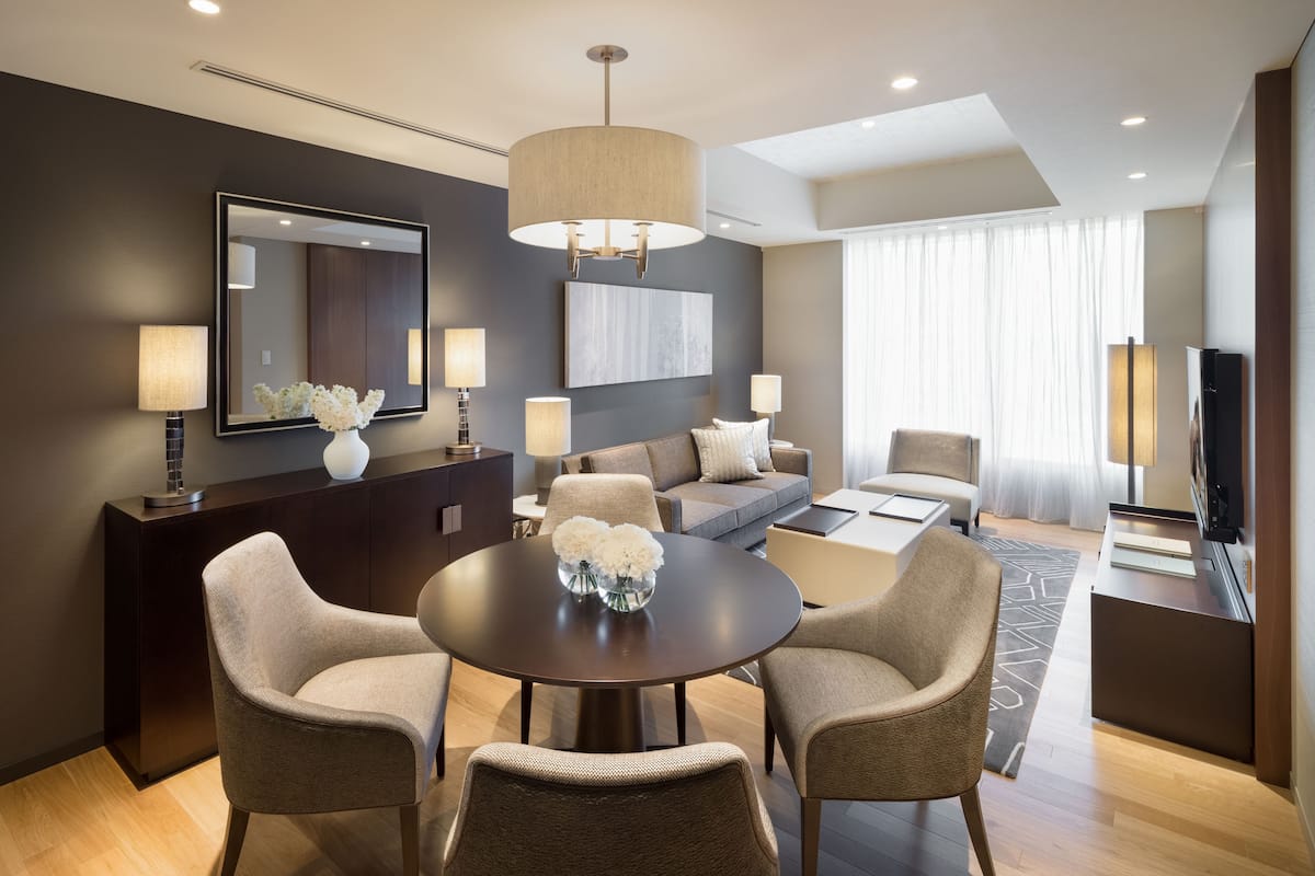 Quarto de hotel moderno com uma mesa de jantar redonda, assentos confortáveis, um espelho grande, televisão de tela plana e uma janela ampla com cortinas leves.