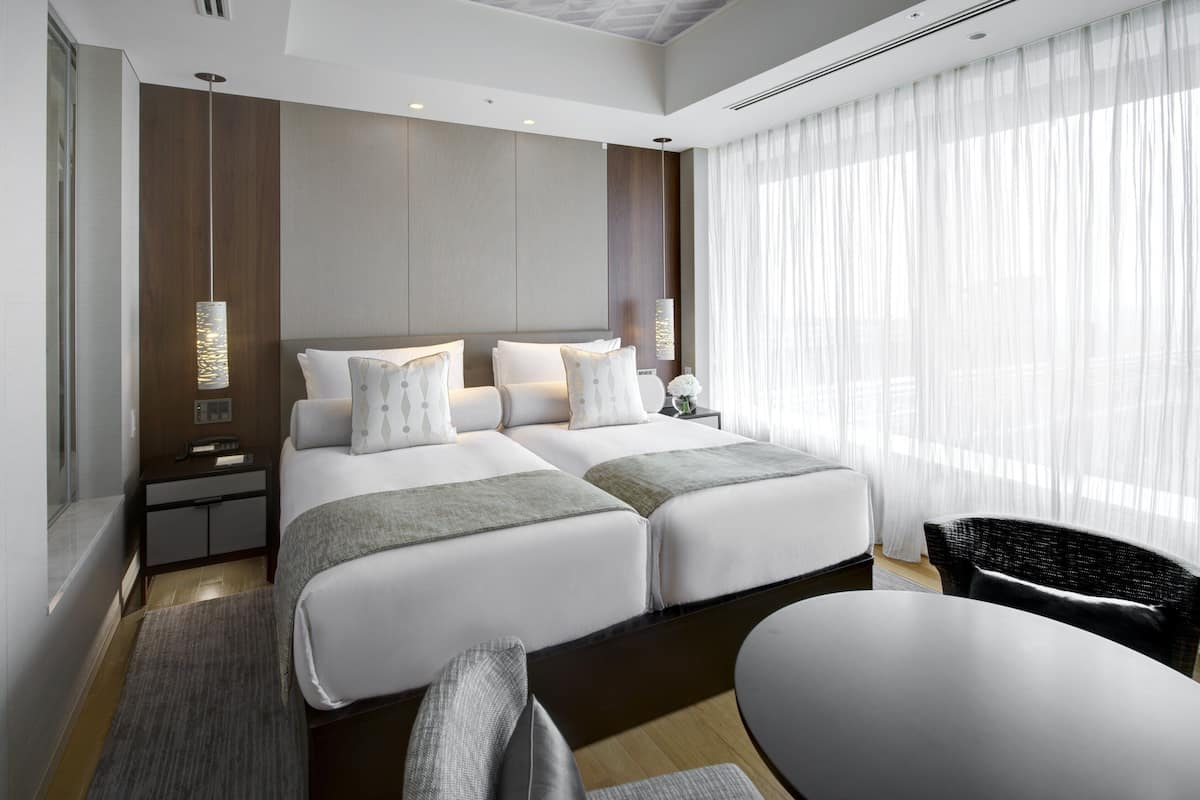 Quarto de hotel moderno com duas camas, uma mesa de jantar e uma janela grande com cortinas leves.