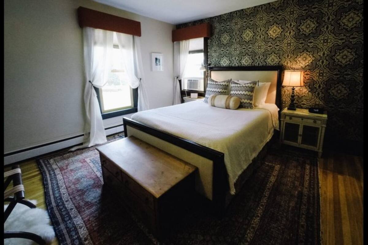 Quarto com uma cama grande, mesa de cabeceira com abajur, um banco e uma cadeira.