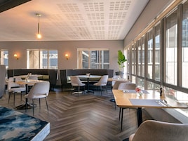 Uma sala de jantar moderna com piso de madeira, mesas, cadeiras e uma grande janela com vista para a cidade.