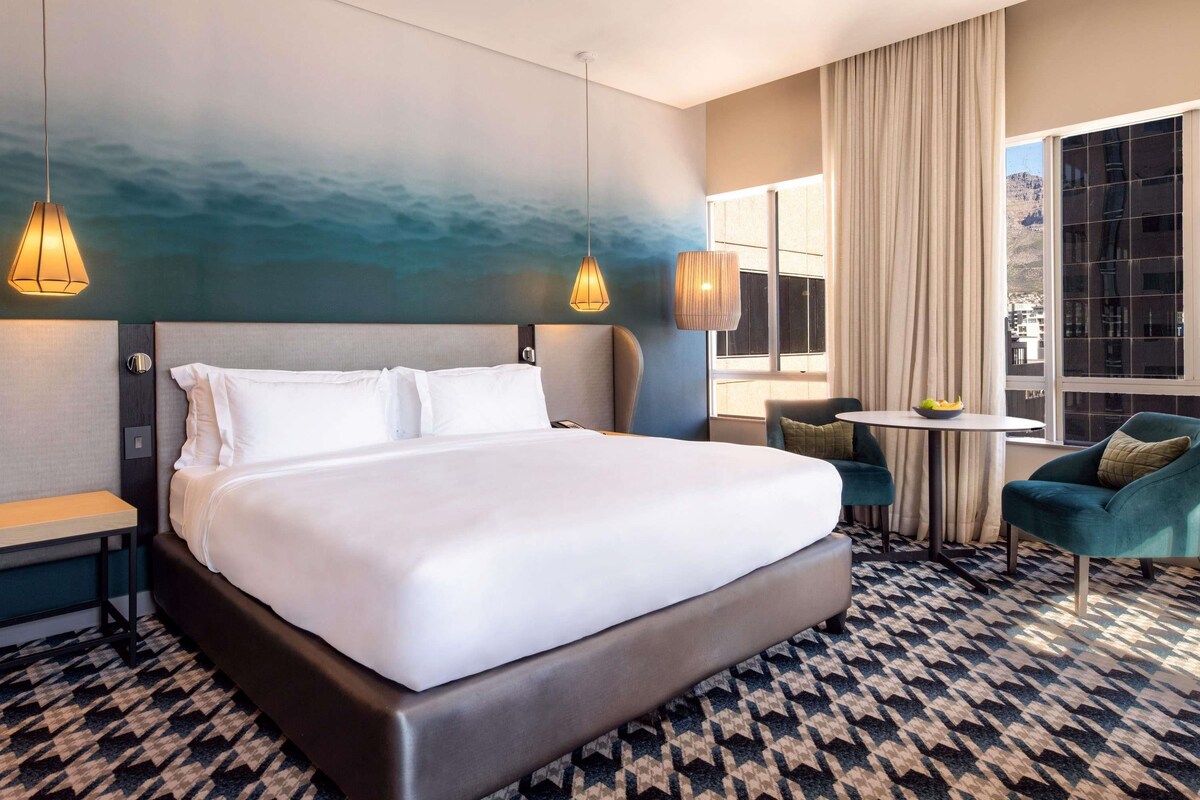Quarto de hotel com uma cama grande, duas mesas de cabeceira, uma mesinha redonda com uma tigela de frutas, uma cadeira e vista da cidade por grandes janelas.