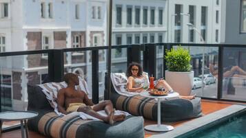 Uma área de piscina no terraço com duas poltronas de tecido, uma mesa com bebidas e um vaso de planta.