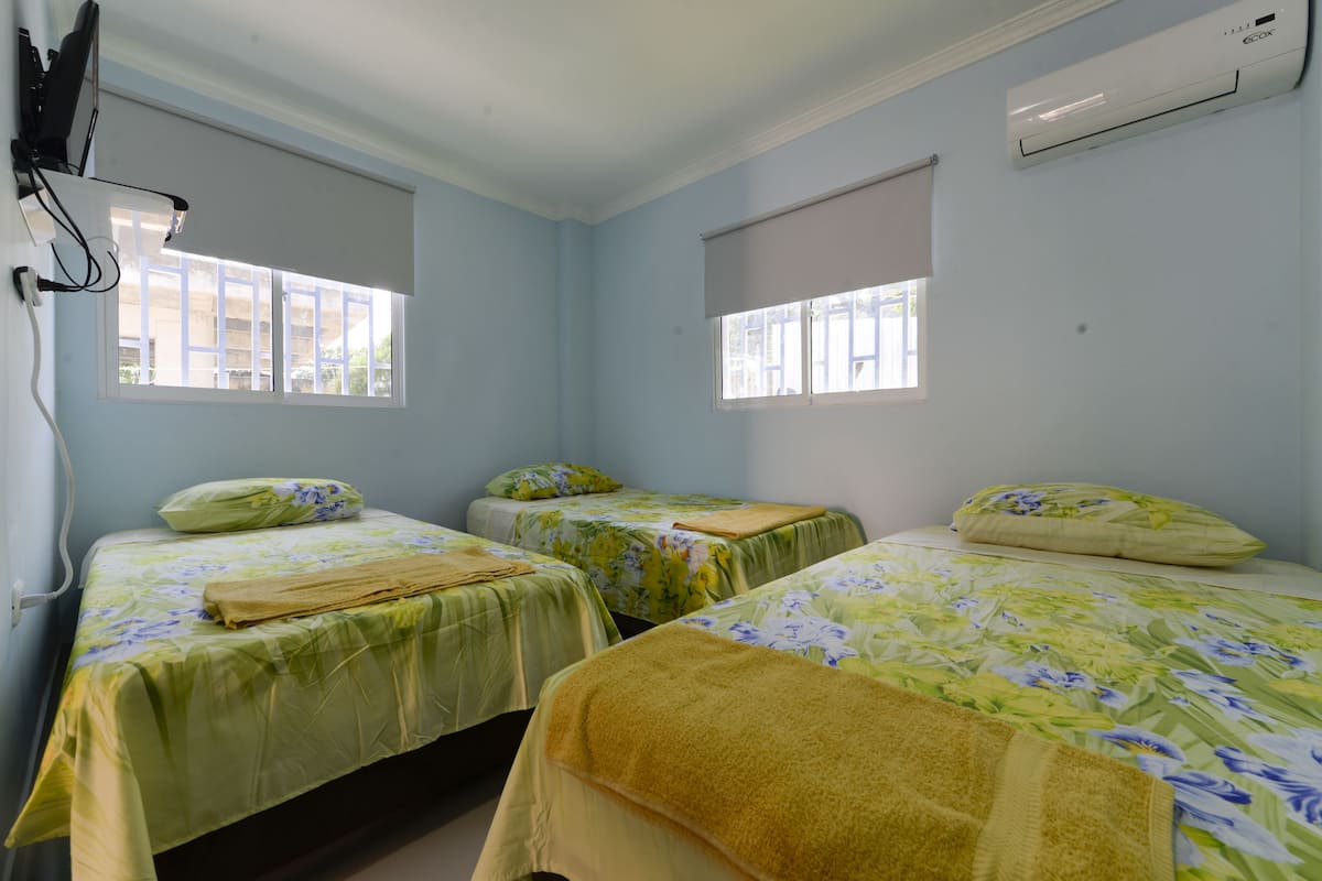 Quarto com três camas, cada uma com roupa de cama e almofadas florais, ar condicionado fixado na parede e uma janela com cortina.