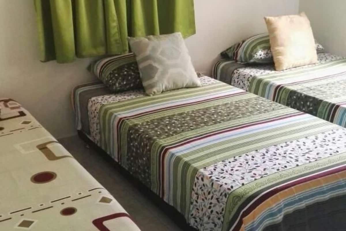 Quarto tipo dormitório com duas camas individuais, cada uma com roupa de cama estampada e almofadas. Uma cortina verde cobre uma janela, e uma barra de metal é visível na parede.