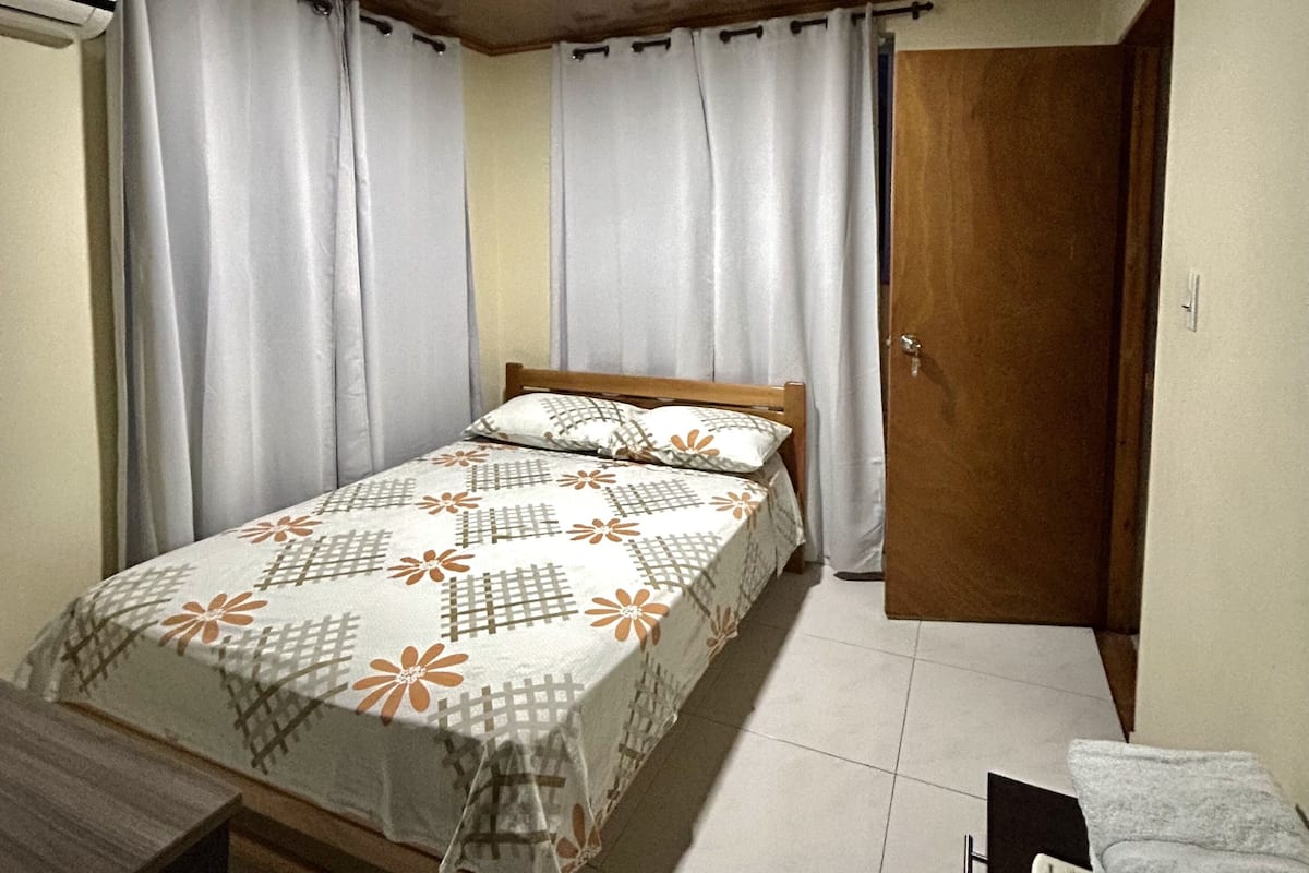 Quarto com cama, cortinas, porta de madeira e uma mesinha com toalha.