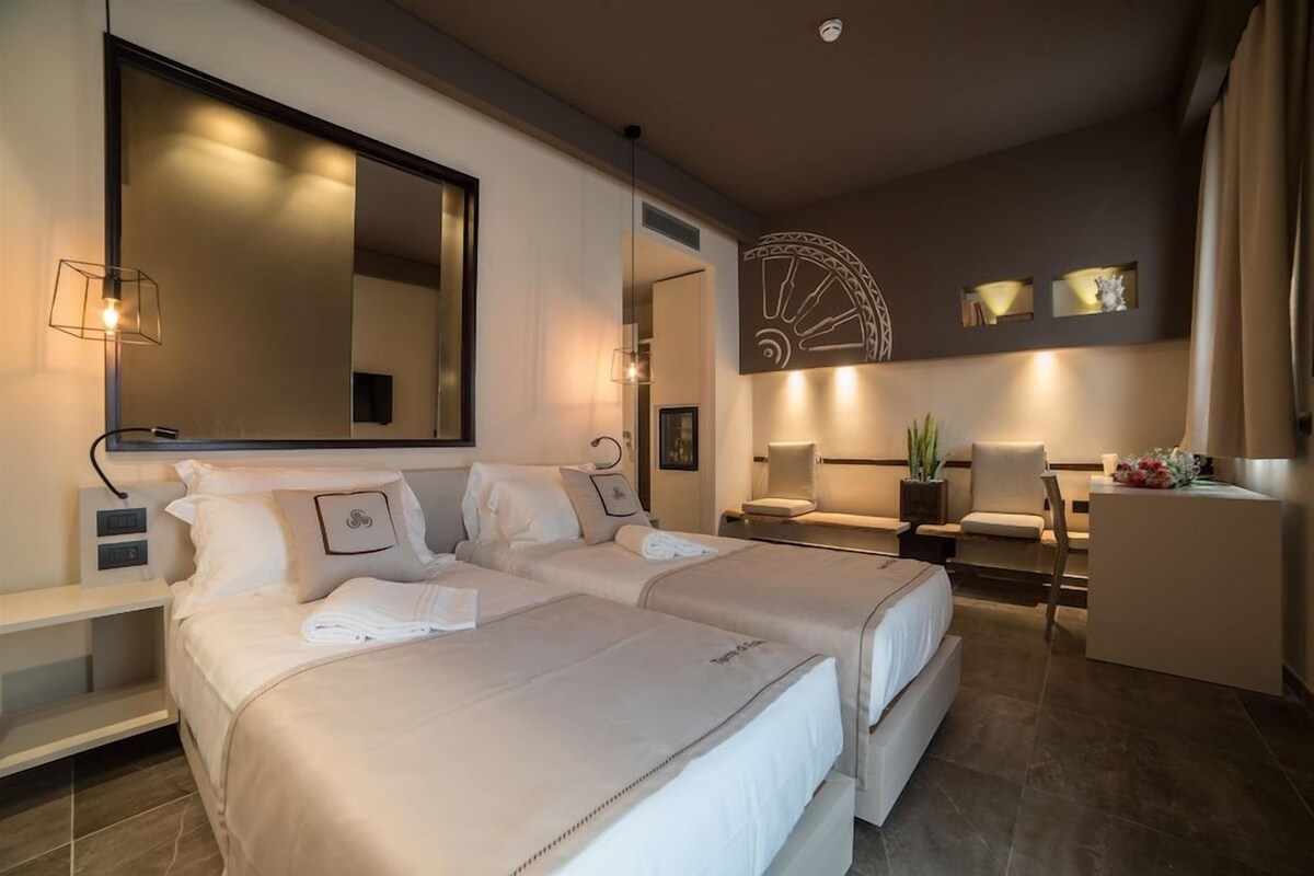 Quarto de hotel com duas camas, uma escrivaninha, uma cadeira, um espelho e um objeto decorativo na parede.