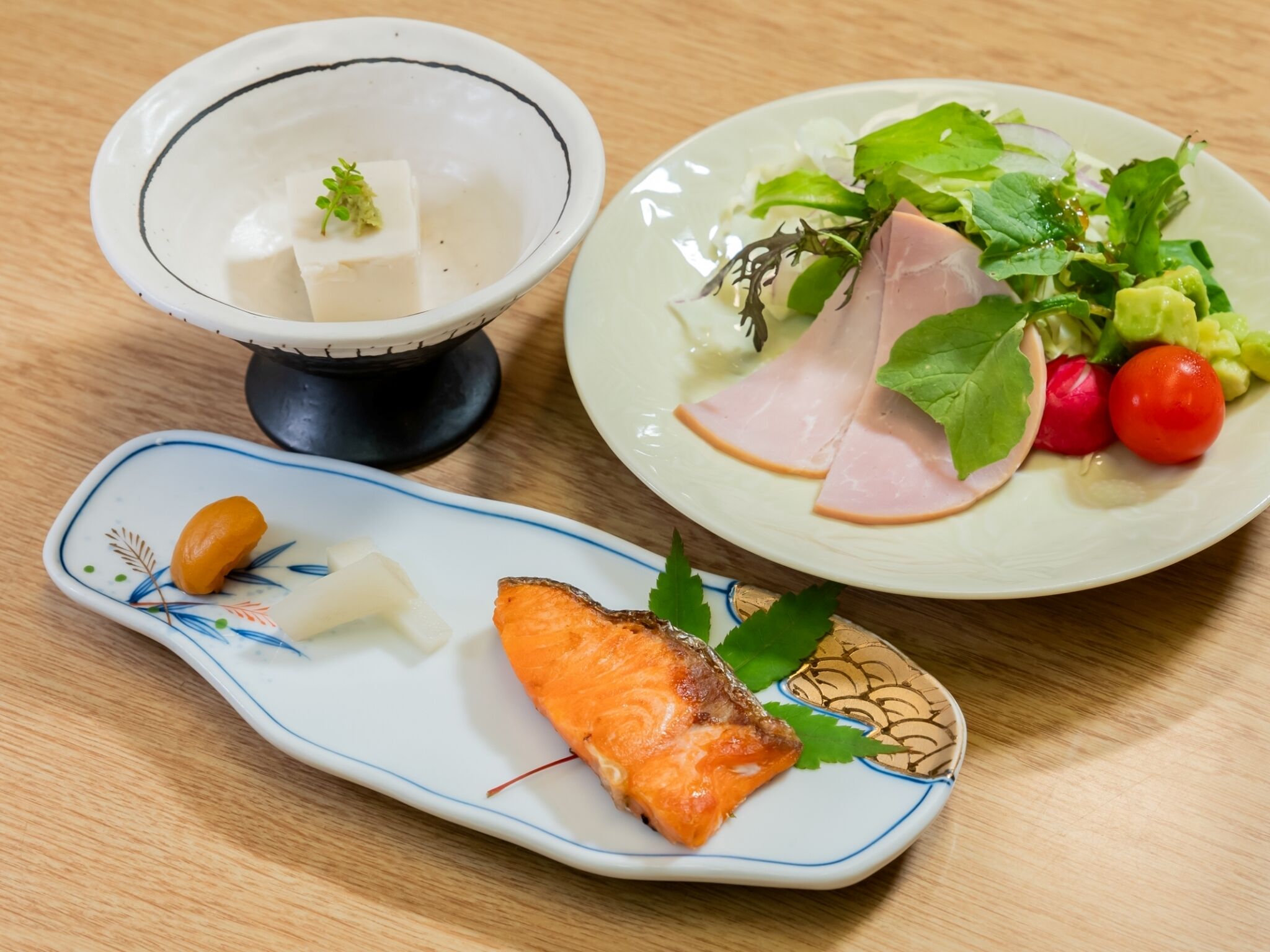 Uma mesa com três pratos: uma tigela de sopa, um prato com salmão e legumes, e um pequeno recipiente com um pedaço de salmão e um tomate cereja.