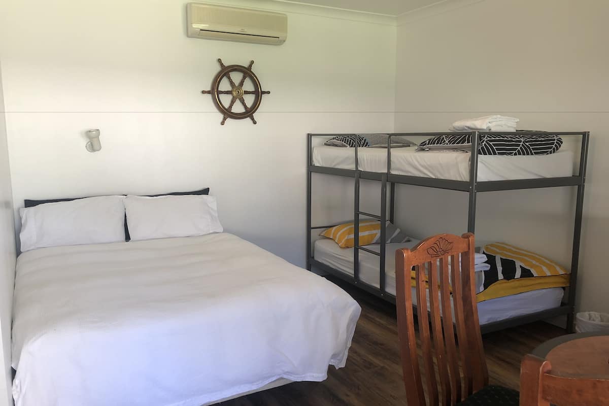 Uma cama de solteiro com roupa de cama branca, uma cama beliche com roupa de cama listrada, uma cadeira de madeira e um leme fixado na parede.
