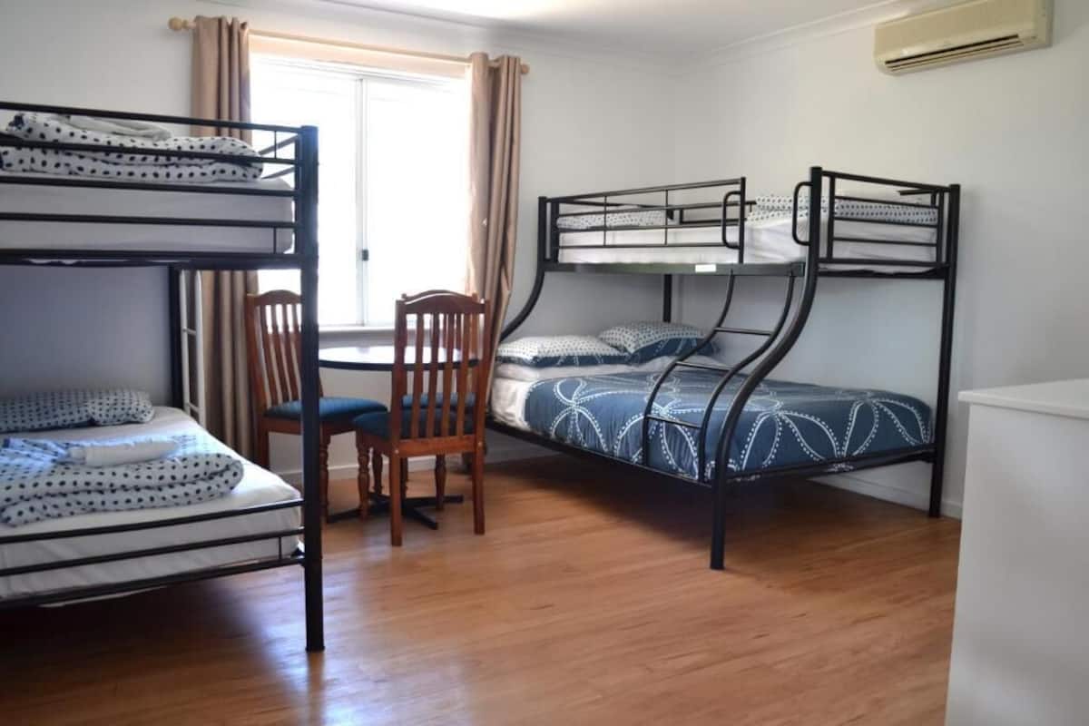 Quarto de dormitório com duas beliches, uma cadeira de madeira, uma escrivaninha e uma janela com cortinas.