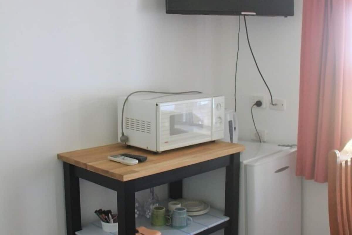 Uma cozinha compacta equipada com micro-ondas, mini-frigorífico, televisão na parede e uma mesa de madeira com um cesto de lixo.