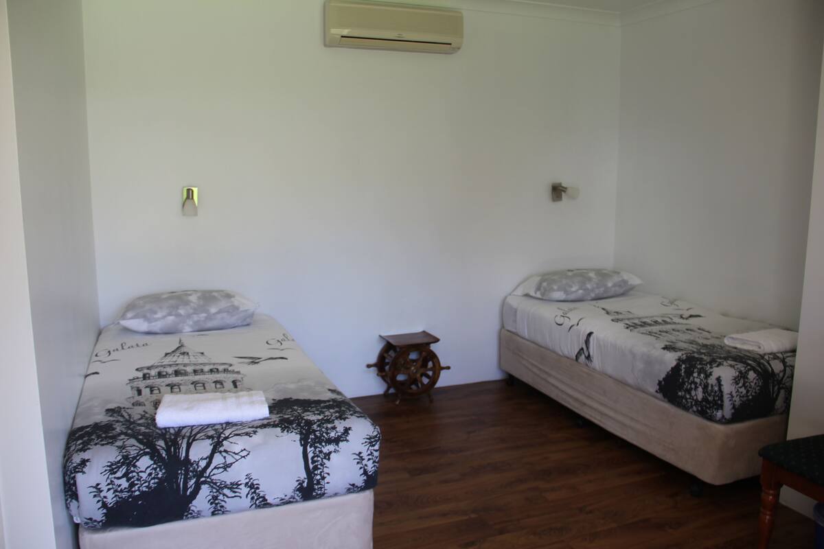 Quarto com duas camas de solteiro, cada uma com um design de colcha diferente, uma pequena mesa de cabeceira de madeira e um aparelho de ar condicionado fixado na parede.