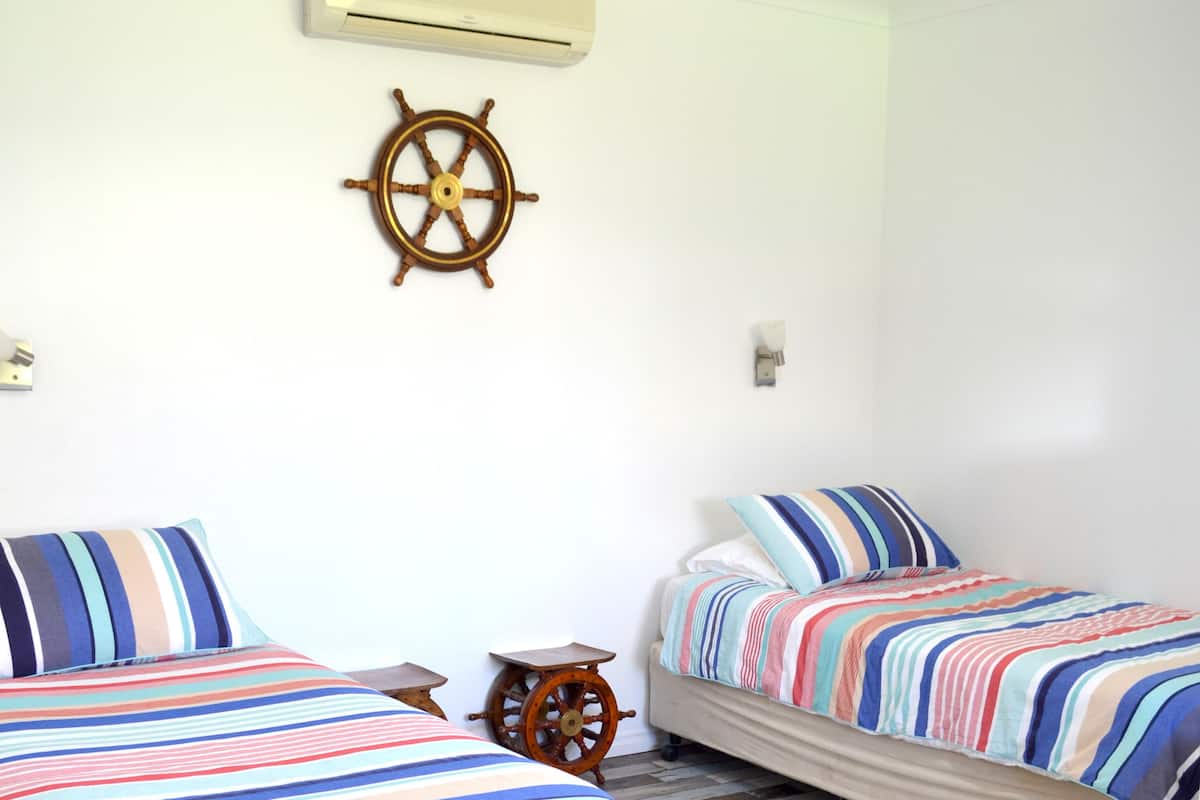 Quarto com duas camas, roupa de cama listrada, um adorno de leme de madeira e um aparelho de ar condicionado branco.