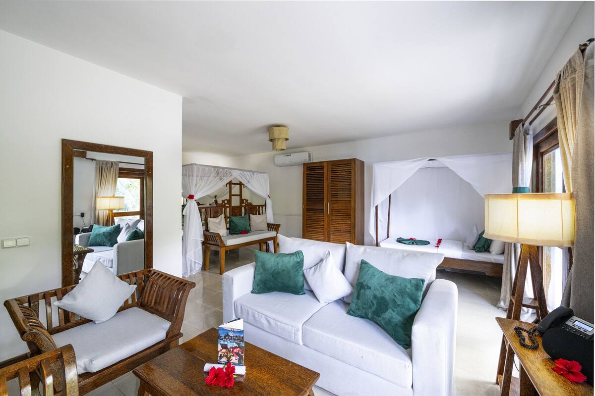 Quarto com um sofá branco, poltronas de madeira, uma mesa de centro em madeira e uma mesa de cabeceira com luminária.