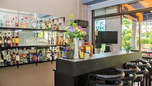 Uma área de bar com diversos frascos de bebidas, copos e um vaso de flores. Há banquetas e uma tela de televisão.