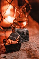 Um copo com líquido âmbar sobre uma base de pedra, uma caixa de petiscos e uma vela acesa ao fundo.