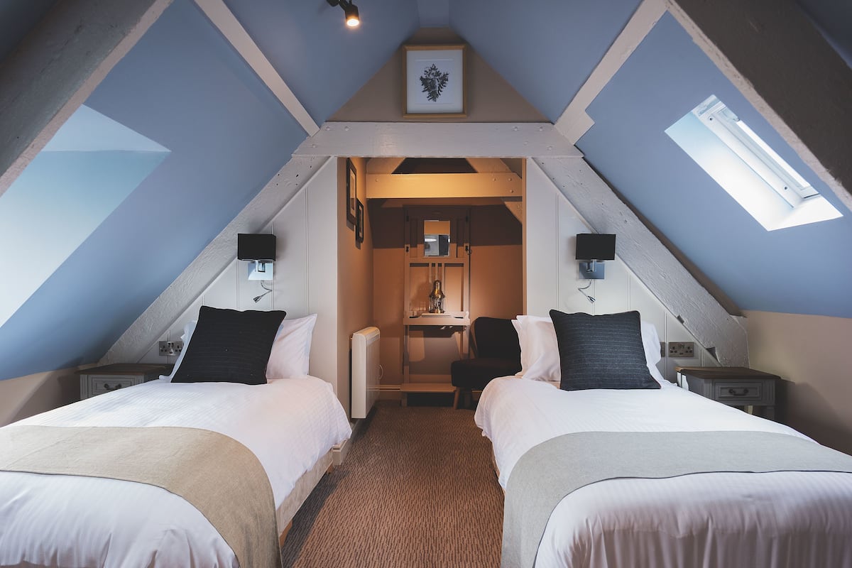 Quarto com duas camas, uma escrivaninha e um claraboia.