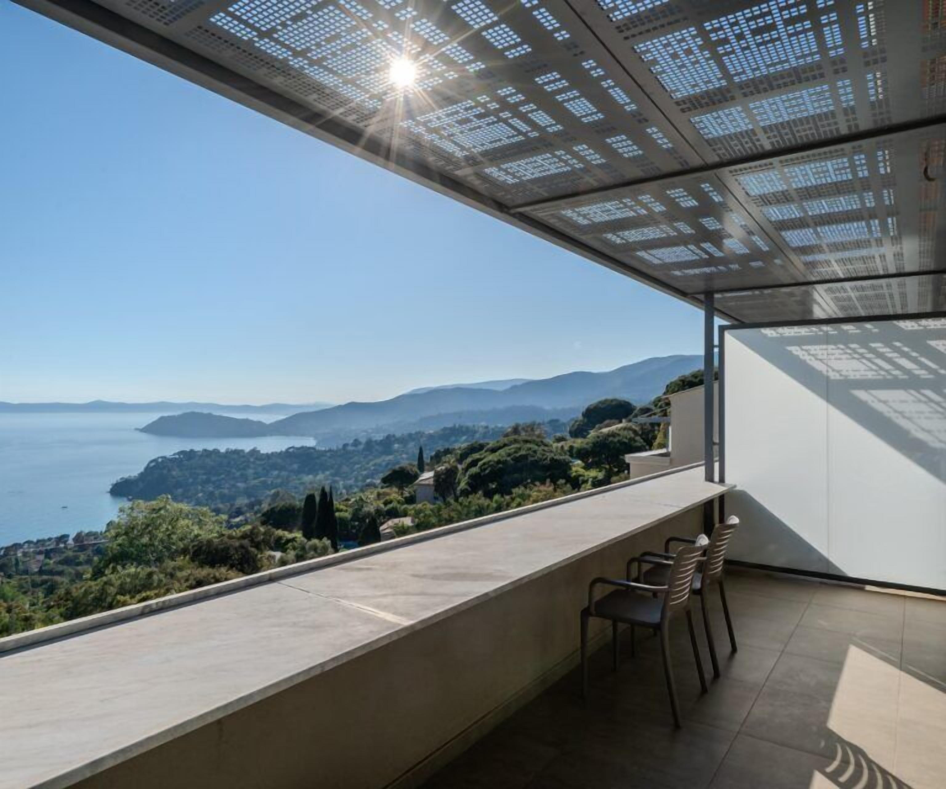Uma varanda com vista para o mar e colinas, uma pérgula e duas cadeiras.