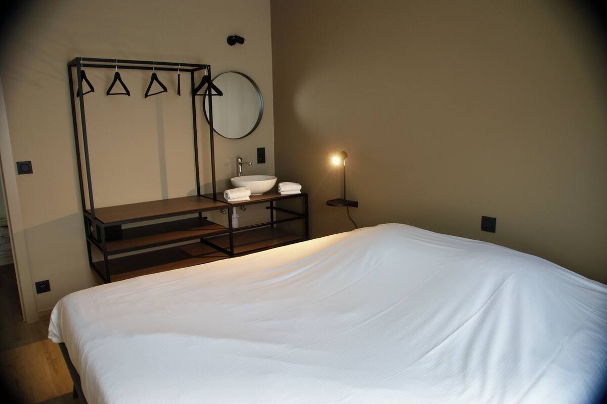 Uma cama bem arrumada com edredom branco, mesa de cabeceira com espelho redondo e pia pequena, além de luminária fixada na parede.