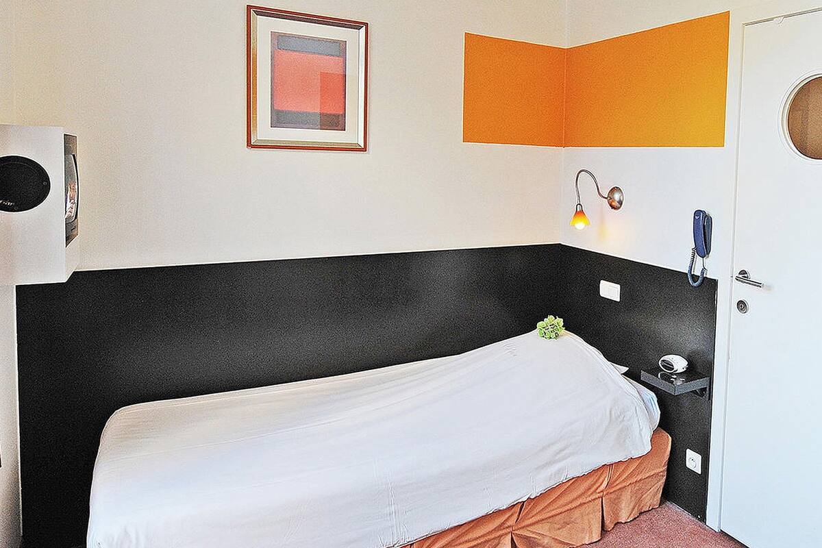 Uma cama de solteiro com colchão branco e cabeceira preta em um quarto com parede branca, um quadro emoldurado e uma mesa de cabeceira com abajur.