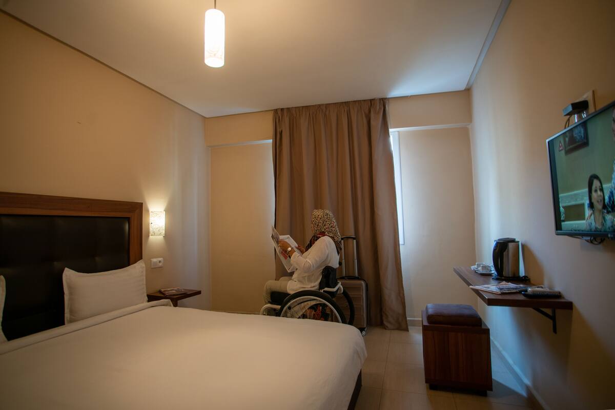 Quarto de hotel com cama, cadeira de rodas, mesa pequena com chaleira e televisão na parede.