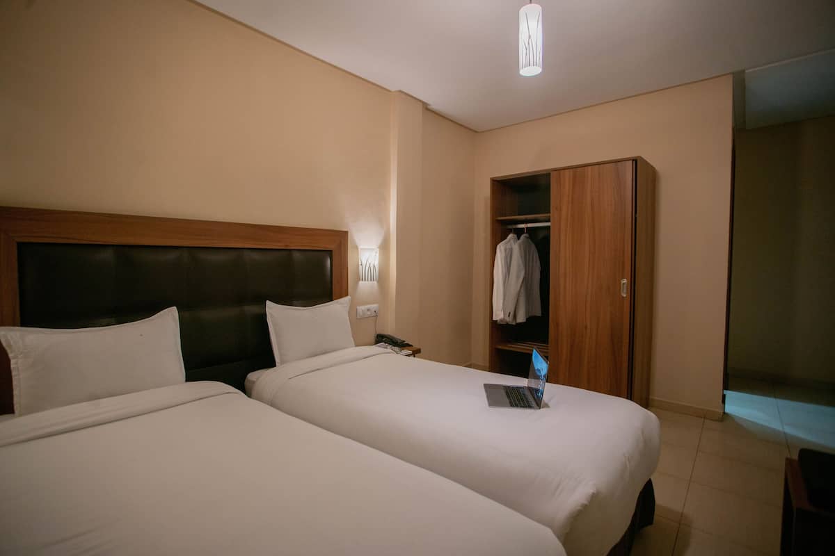 Quarto de hotel com duas camas, um guarda-roupa de madeira e uma pequena mesa de escritório com um laptop.