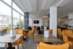Restaurante moderno com cadeiras e mesas amarelas, janelas amplas e uma televisão de tela plana na parede.