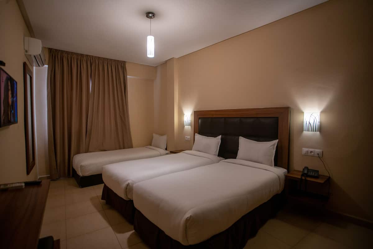 Quarto de hotel com duas camas, cabeceira de madeira, luminárias de cabeceira, ar condicionado e telefone num criado-mudo.